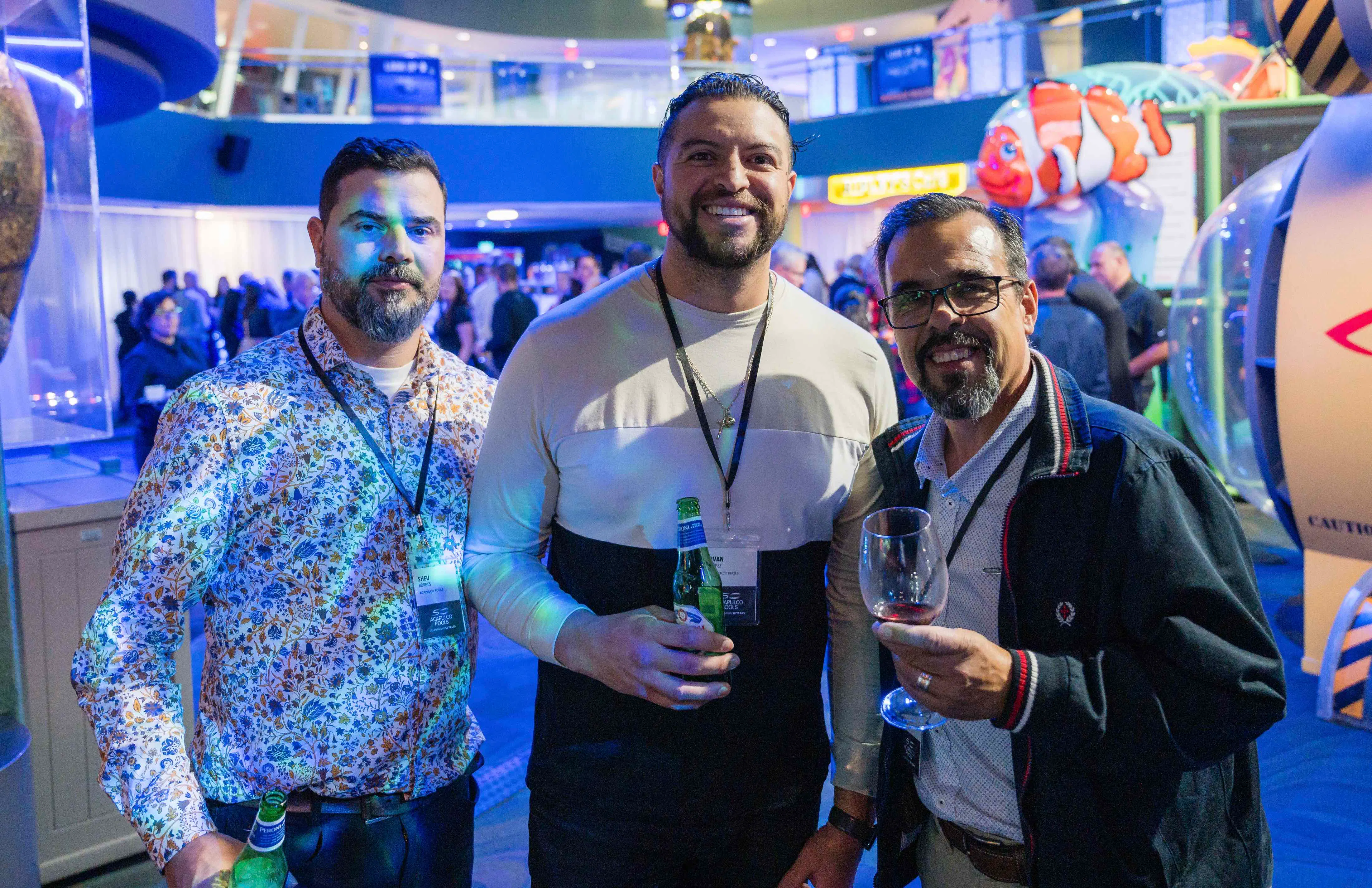 Three men at an event holding drinks, smiling in a brightly lit venue.