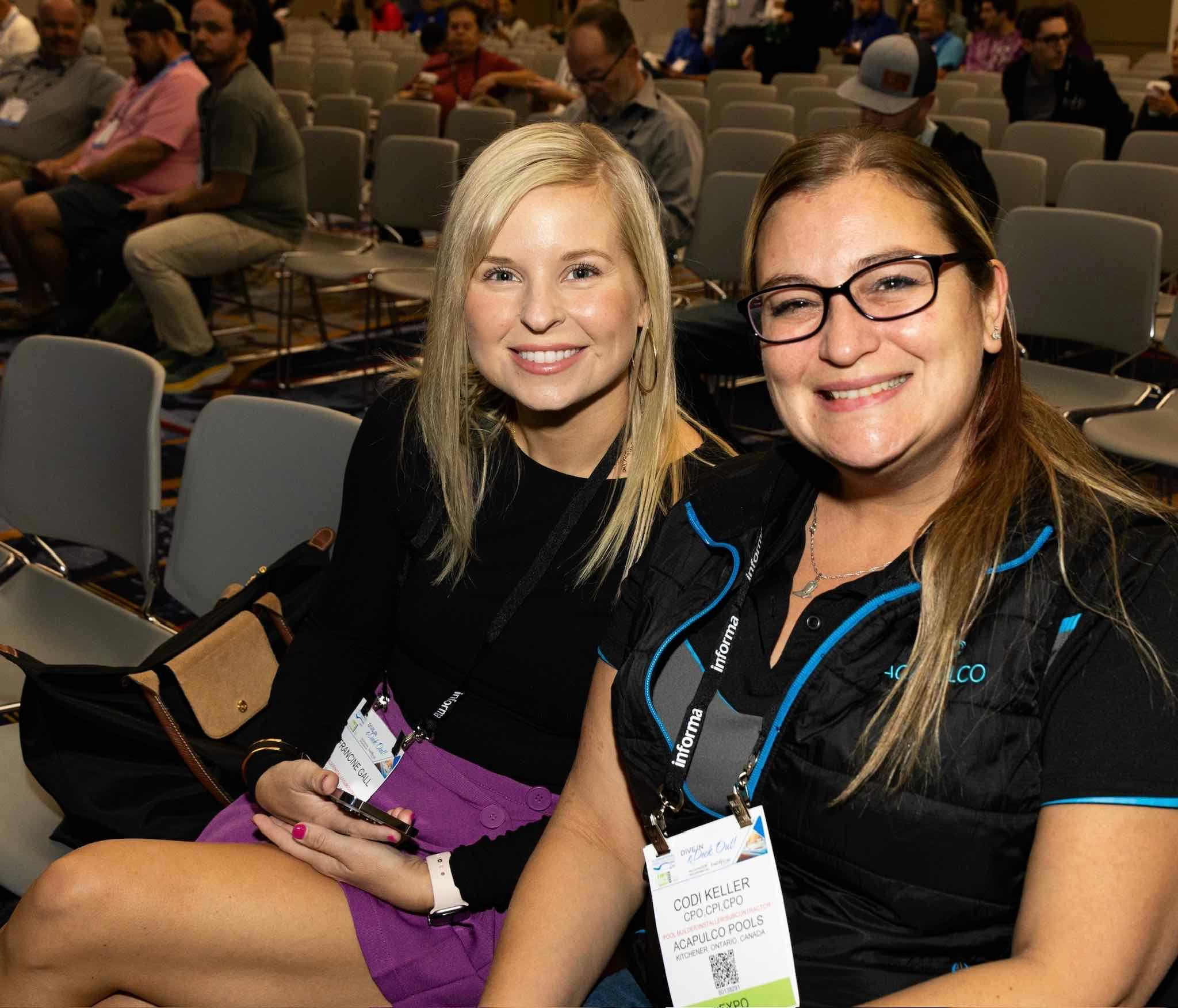 Two women smiling while sitting at a conference, one holding a badge ...
