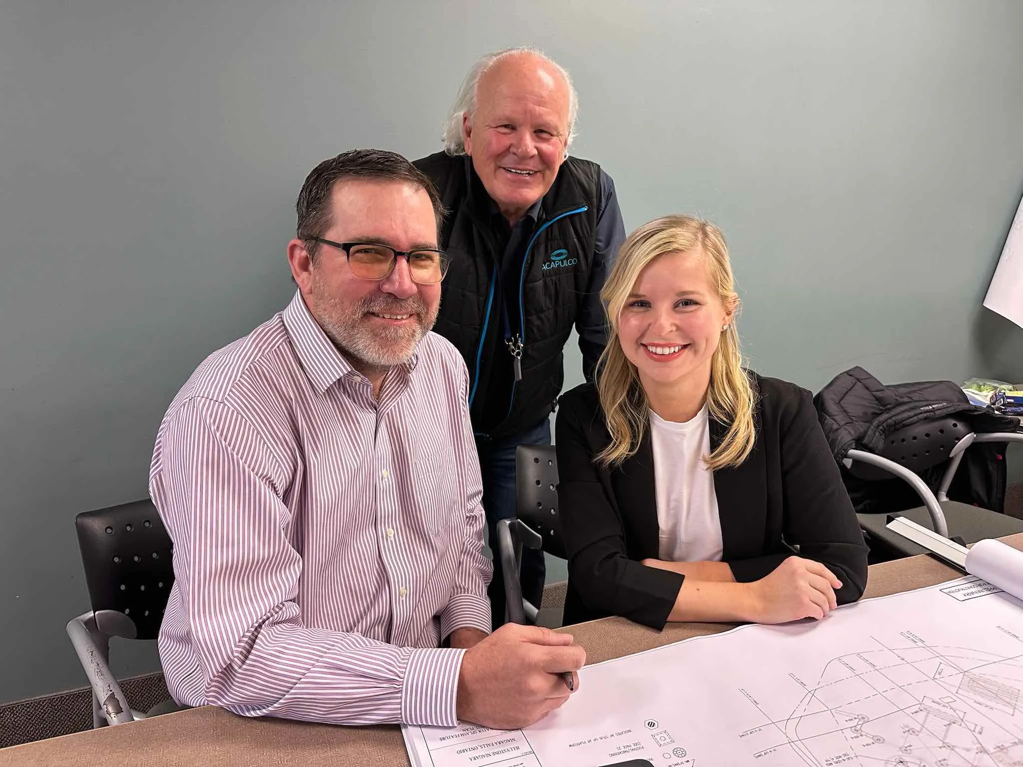 Three people gathered at a table reviewing pool design plans, smiling ...
