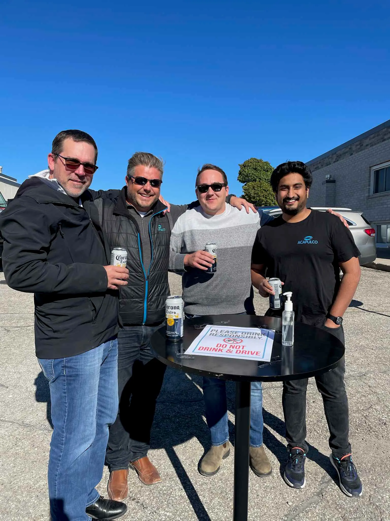 Four men holding drinks, posing outdoors near a table with a “Drink ...