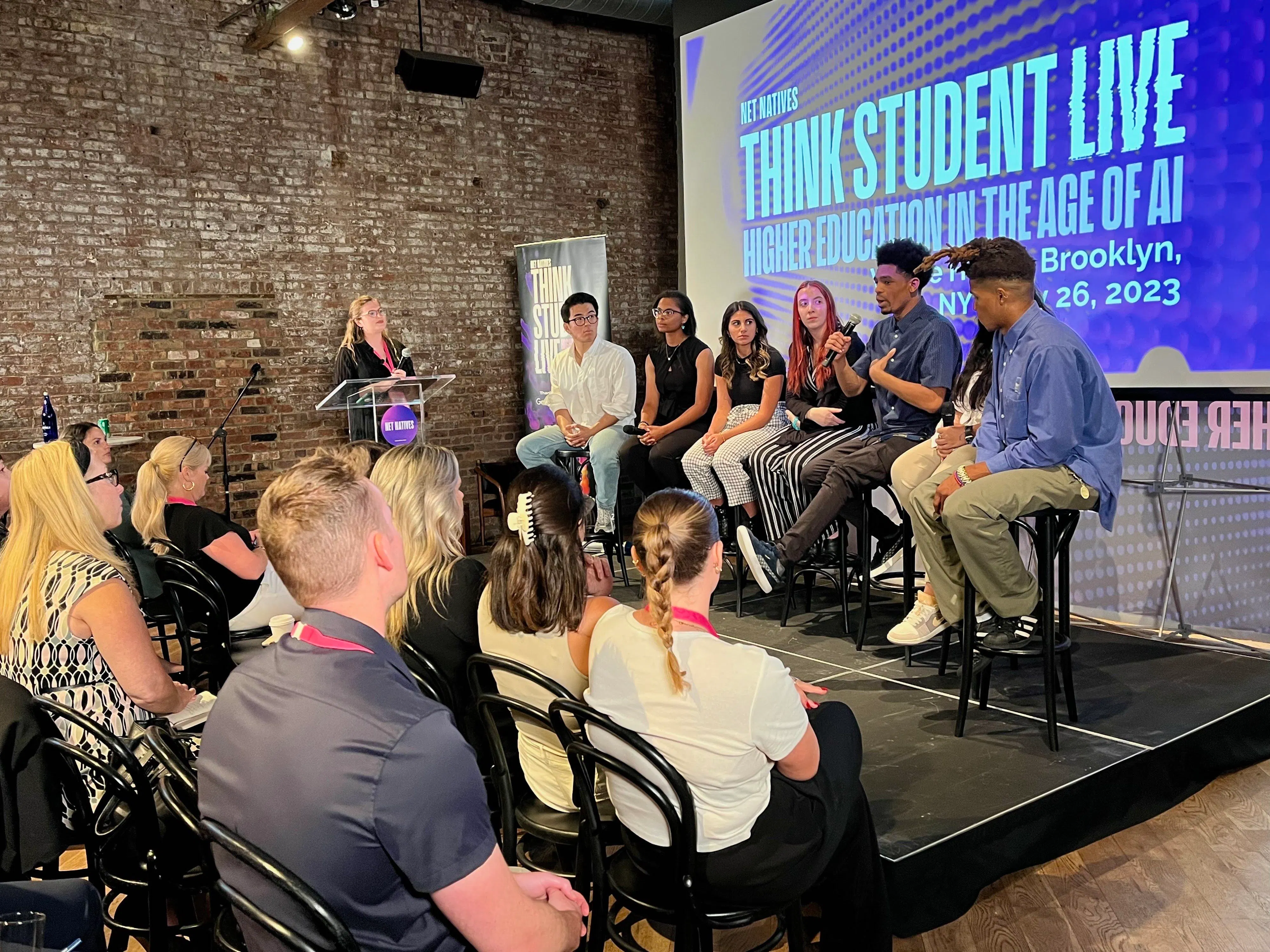 Seven students sit on stools facing an audience, while one woman moderates.