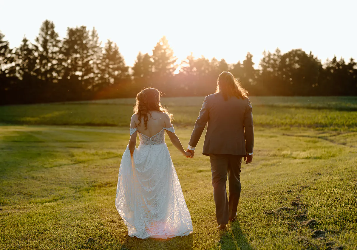 Bride and groom walking hand in hand through a grassy field at sunset.